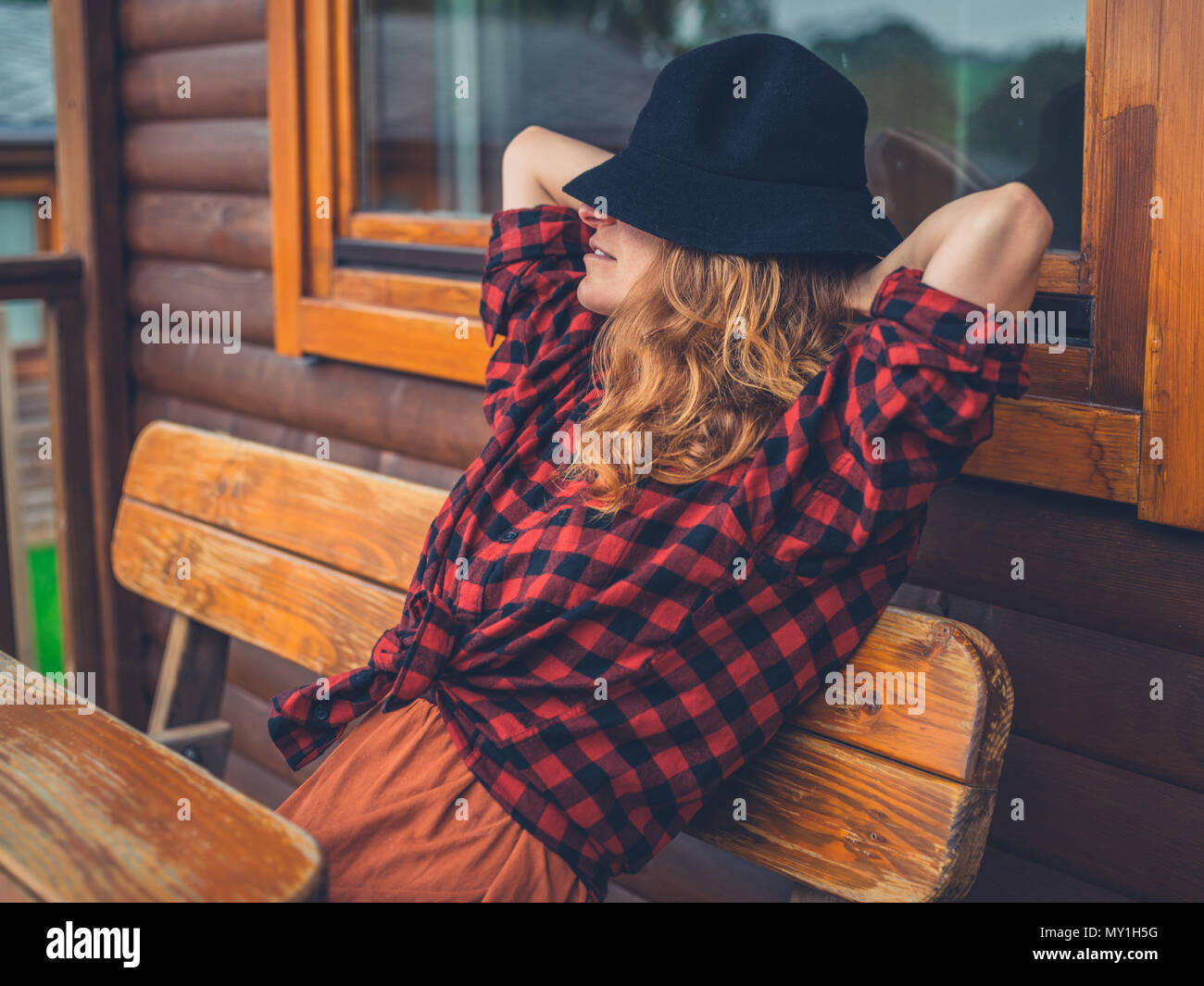 A young woman wearing a hat is relaxing on the porch of a log cabin ...
