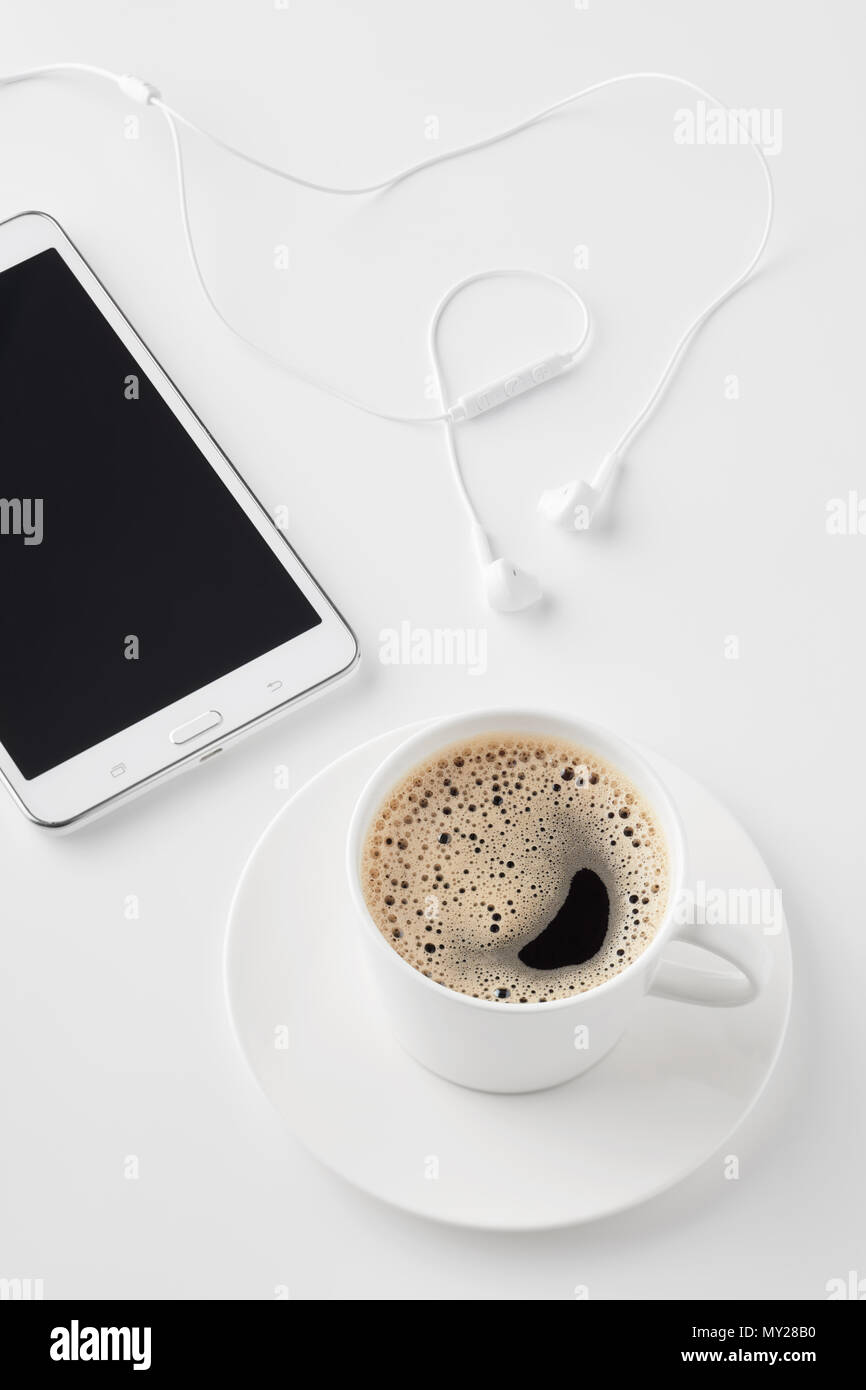 Coffee with foam in a white cup, earphones, and tablet on white table ...