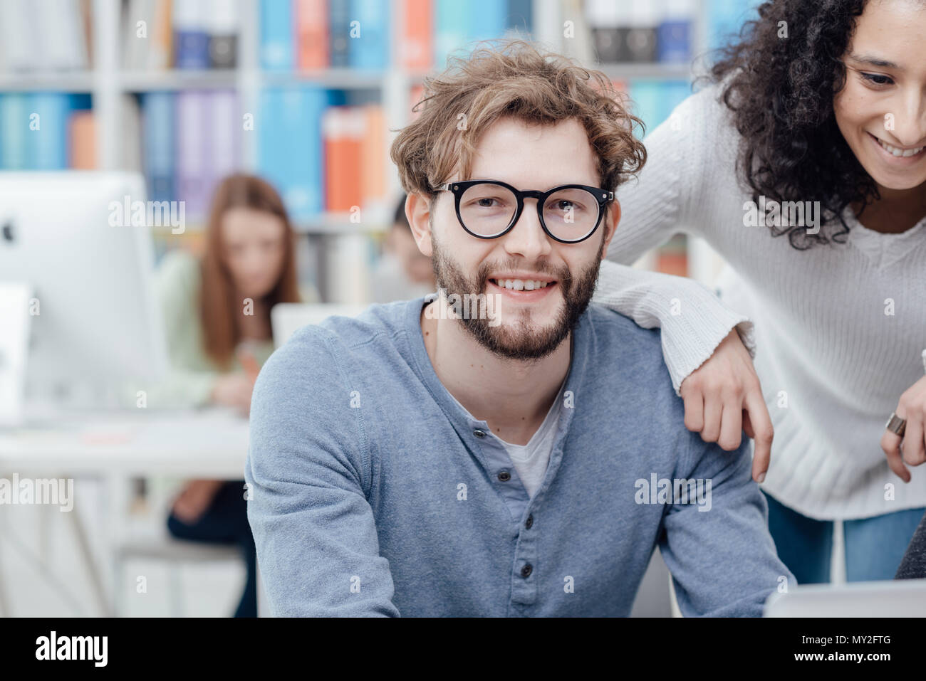 Young smiling hipster man sitting at desk, his colleague is leaning on ...