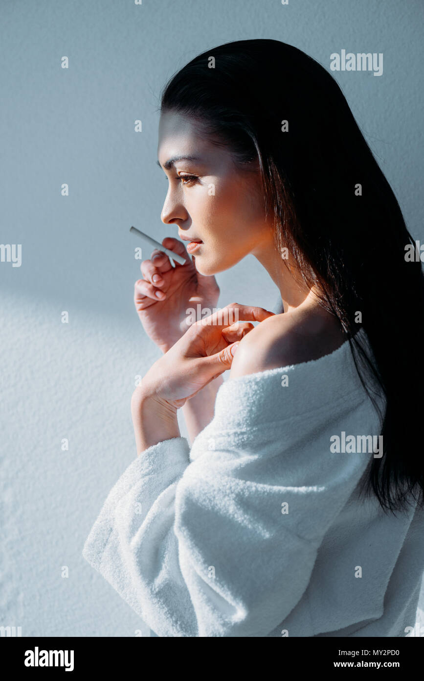 Side view shot of young woman in white bathrobe smoking a cigarette ...