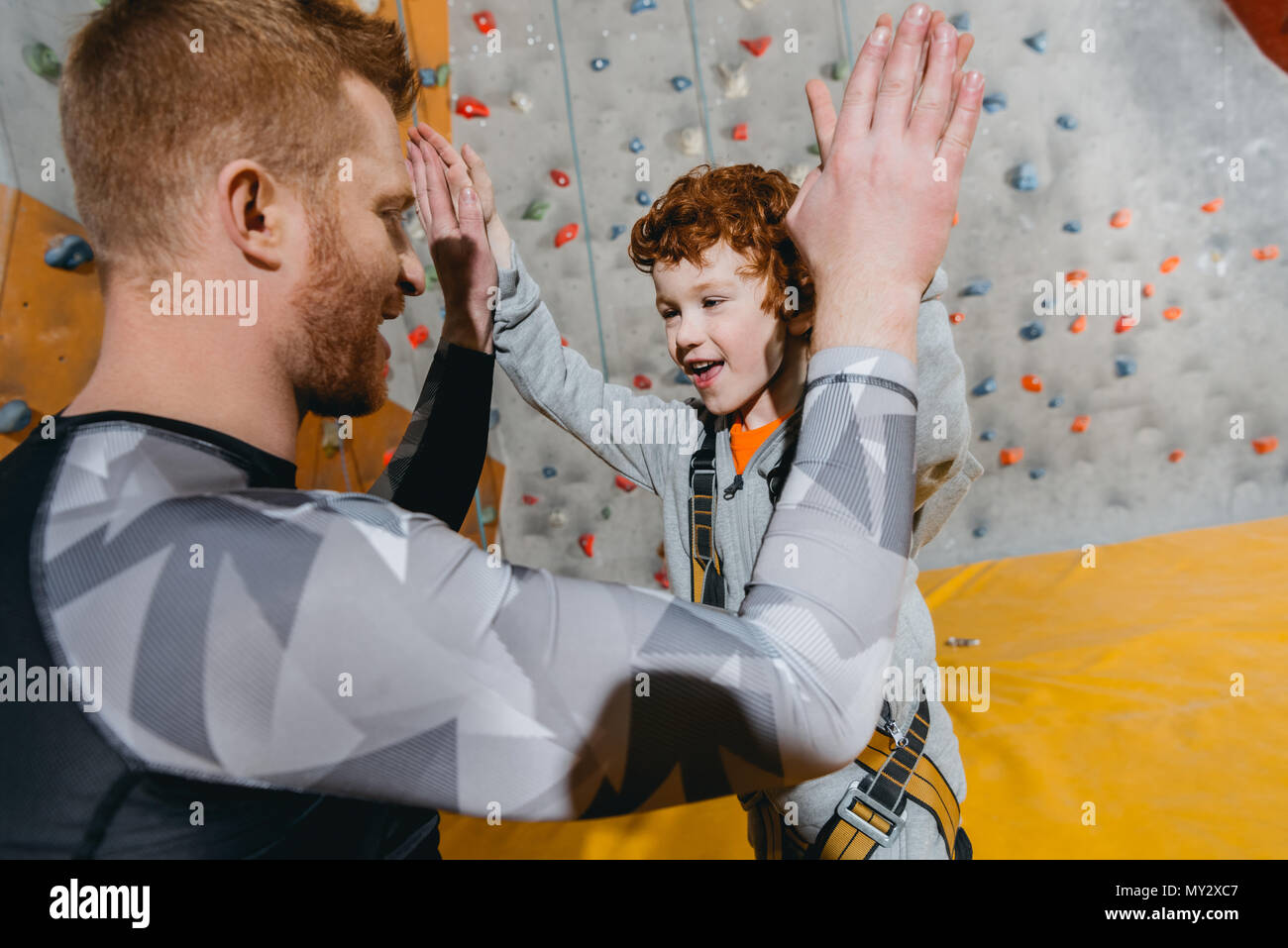 Closeup shot of little boy in harness high-fiving his dad with climbing ...