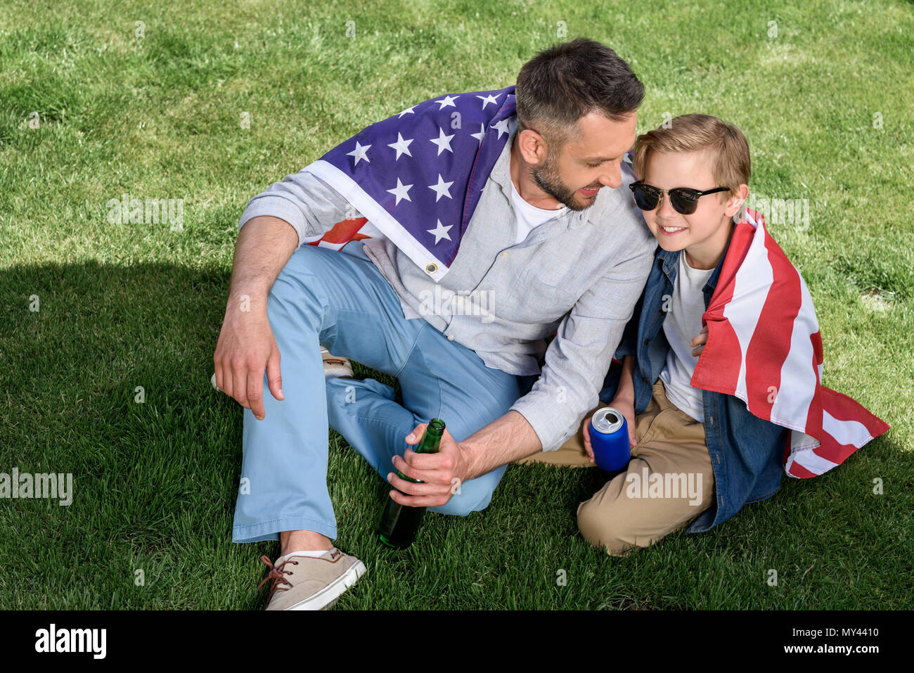 father and son sitting on grass with us flag and holding beer and soda ...