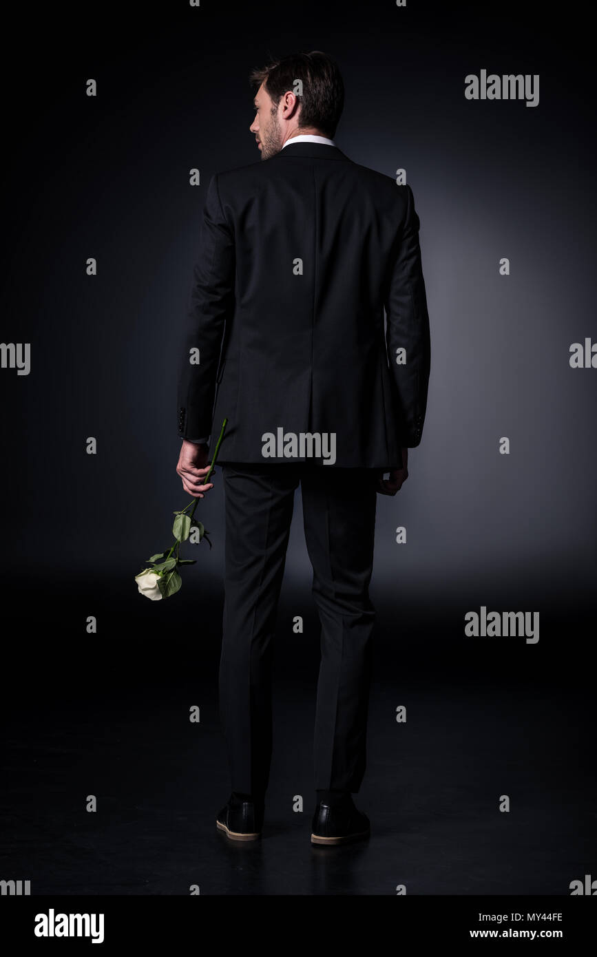 back view of man in formal wear holding white rose isolated on black ...