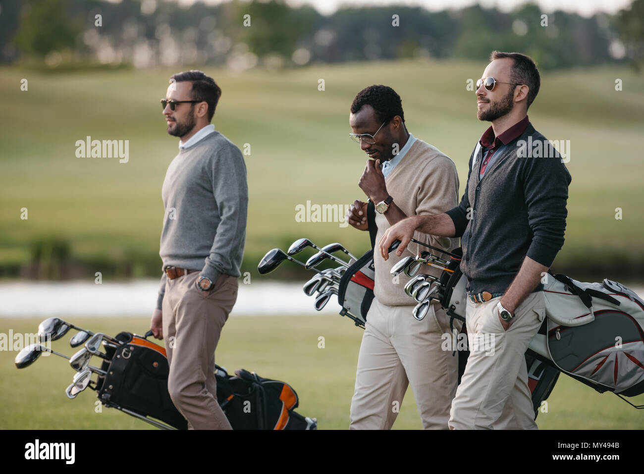 multiethnic golfers holding bags with golf clubs and walking on golf ...