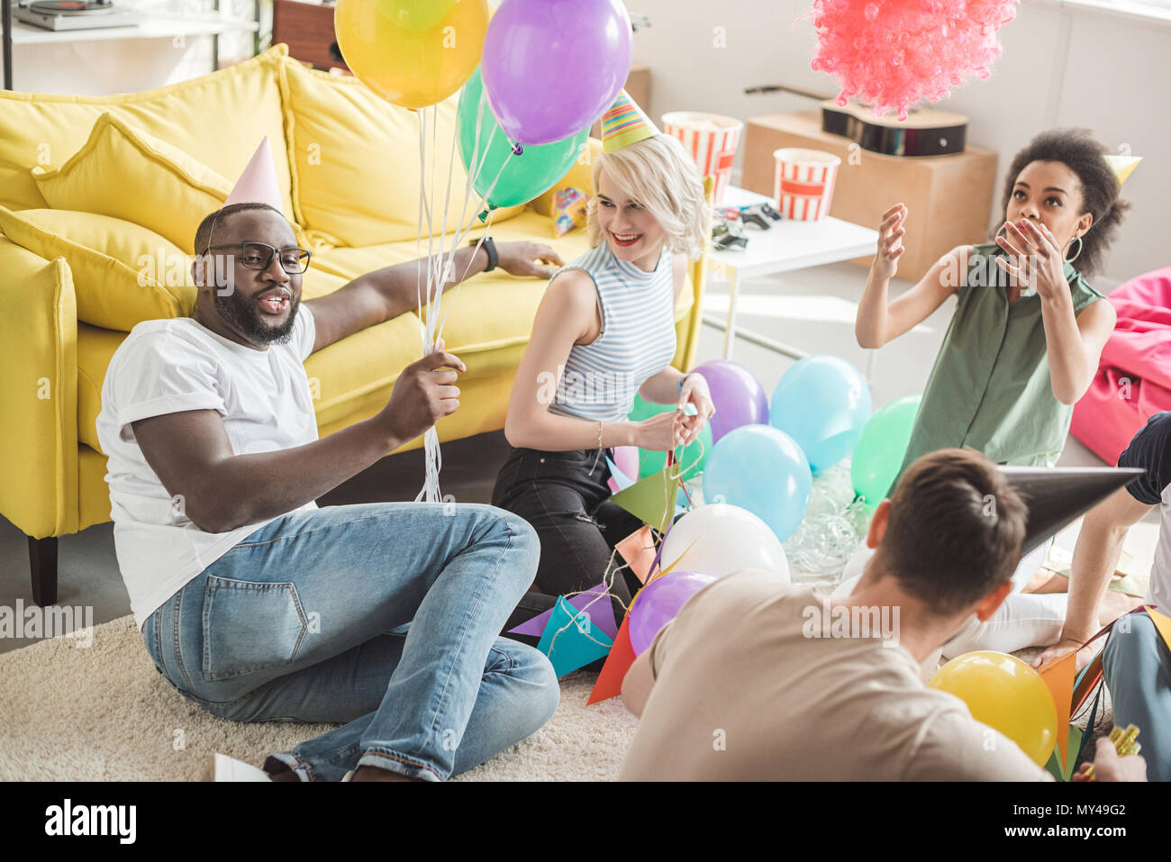multicultural friends in party hats sitting on floor with balloons in ...