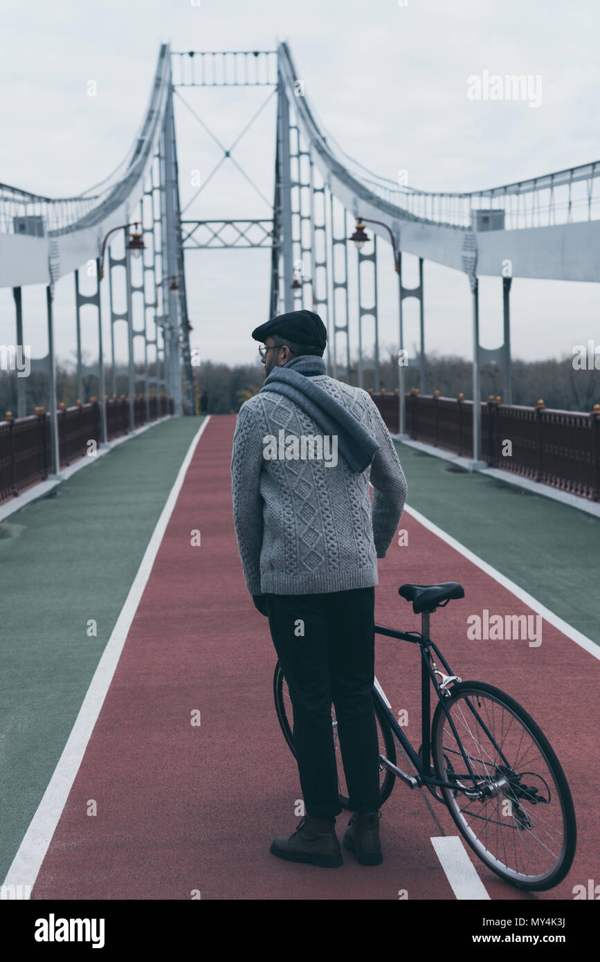 back view of stylish man with bicycle standing on pedestrian bridge ...