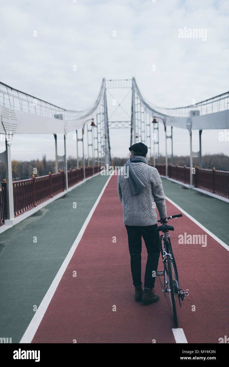 back view of stylish man with bicycle standing on pedestrian bridge ...