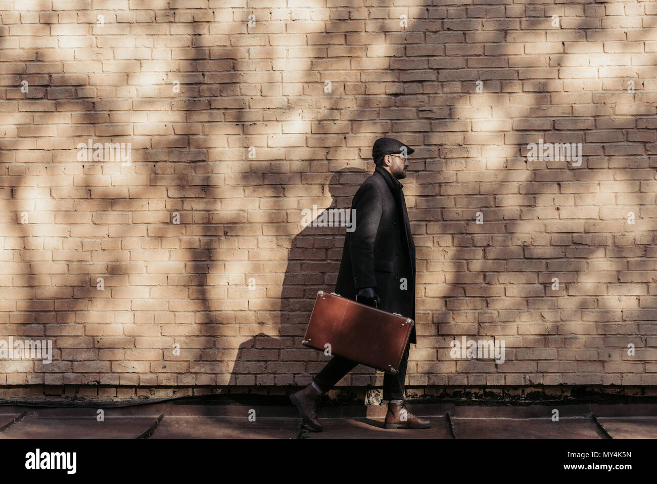 side view of adult man with suitcase walking in front of brick wall ...