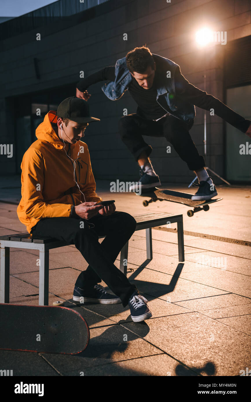 skateboarder jumping over bench while man sitting on it and listening ...