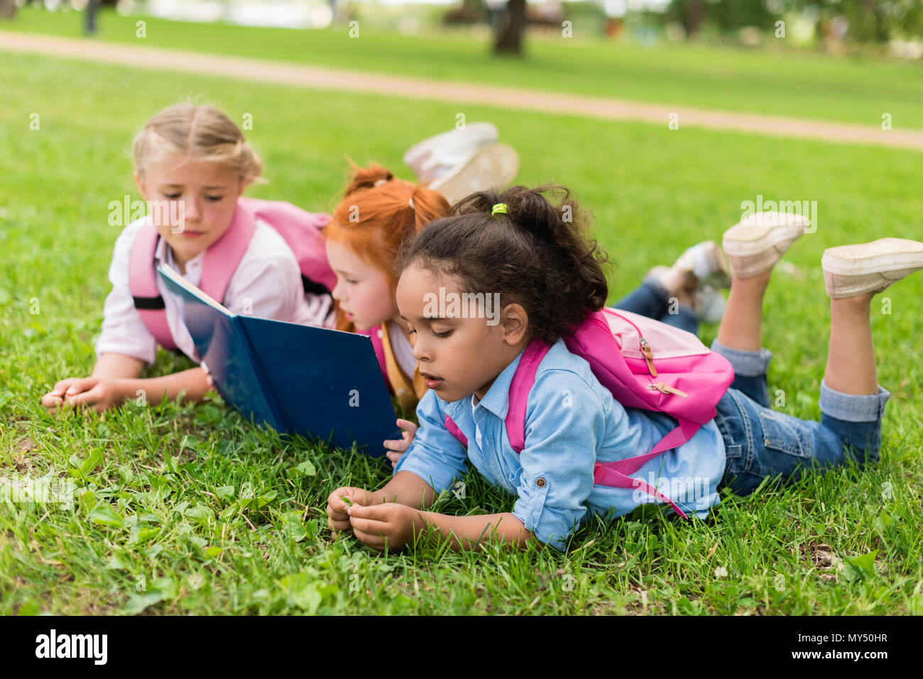 cute little multiethnic schoolgirls reading book together on grass ...