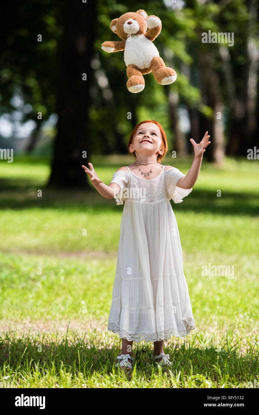 happy little girl in white dress throwing teddy bear while standing in ...