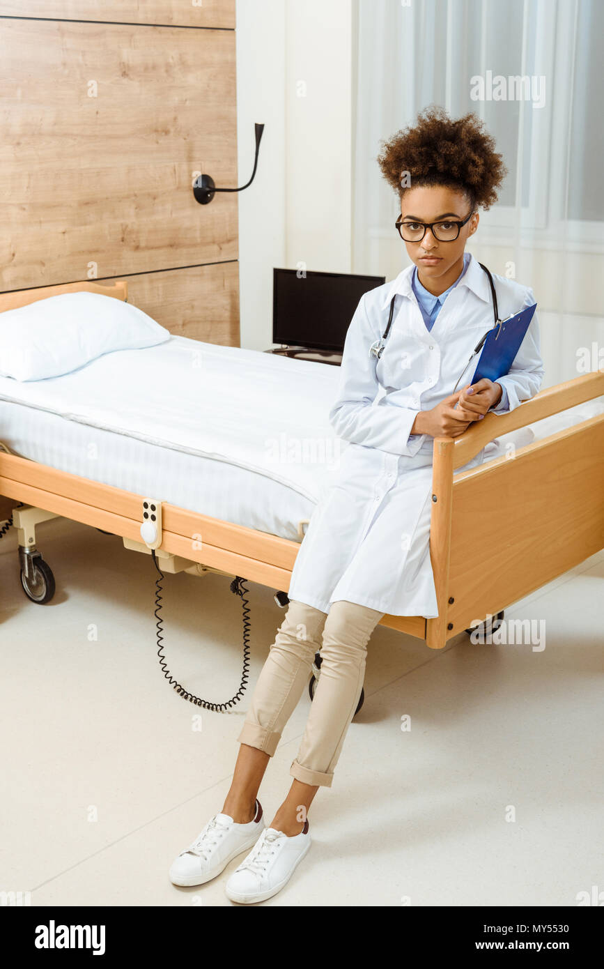 Young african-american doctor in glasses holding clipboard and sitting ...
