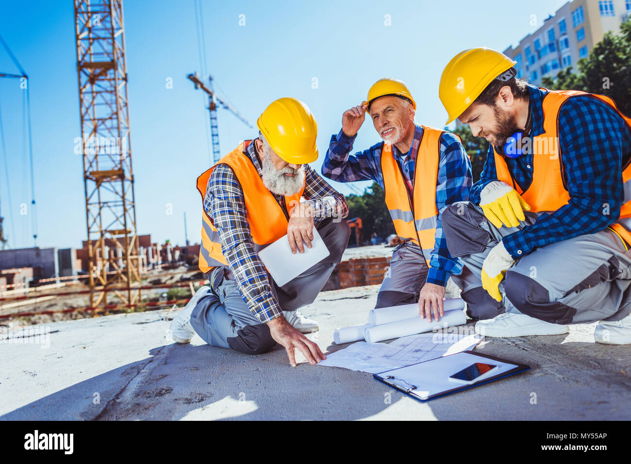 Three construction workers in uniform sitting on concrete at construction site, examining ...