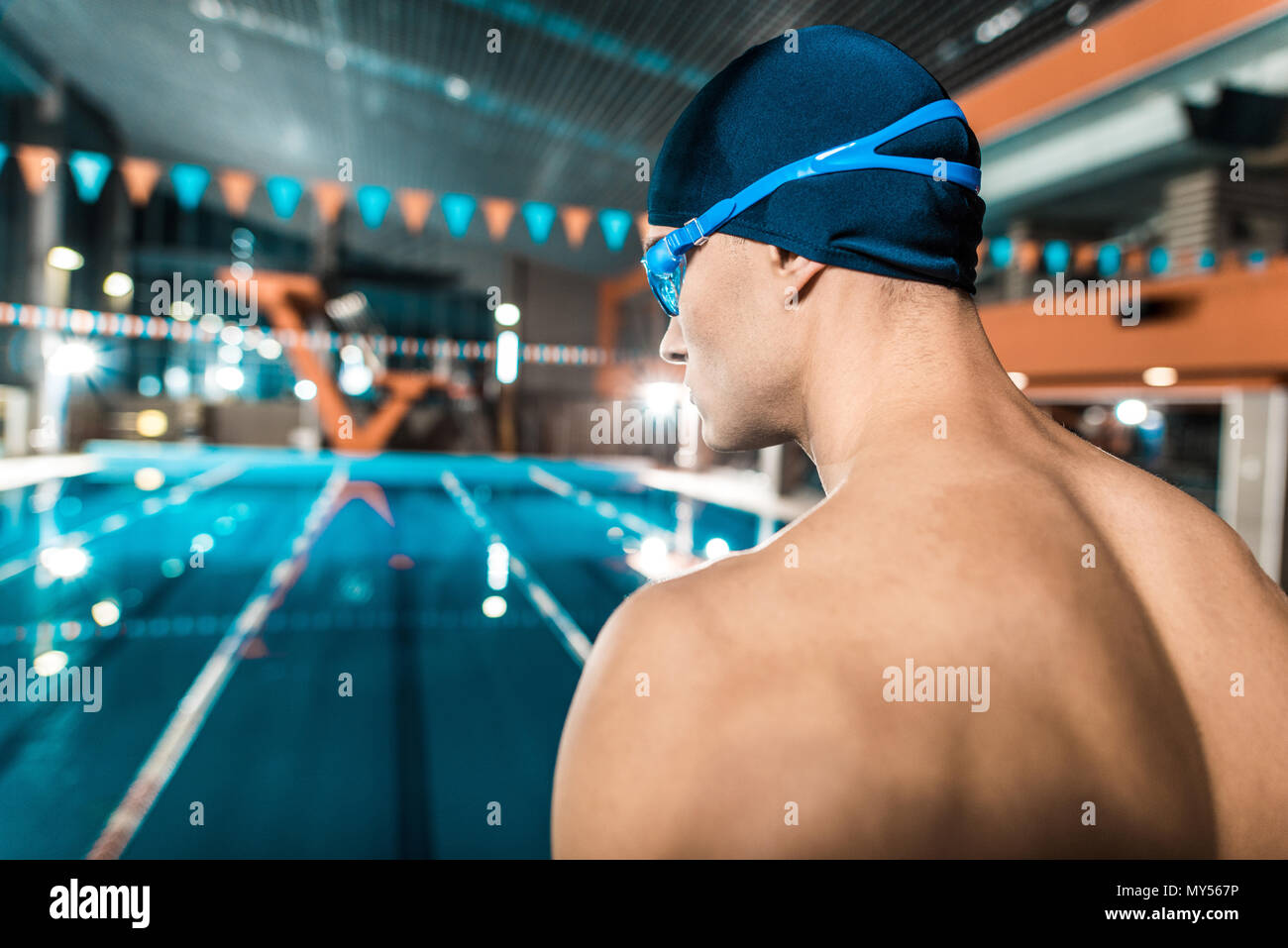 back view of muscular swimmer in swimming cap and goggles standing at ...