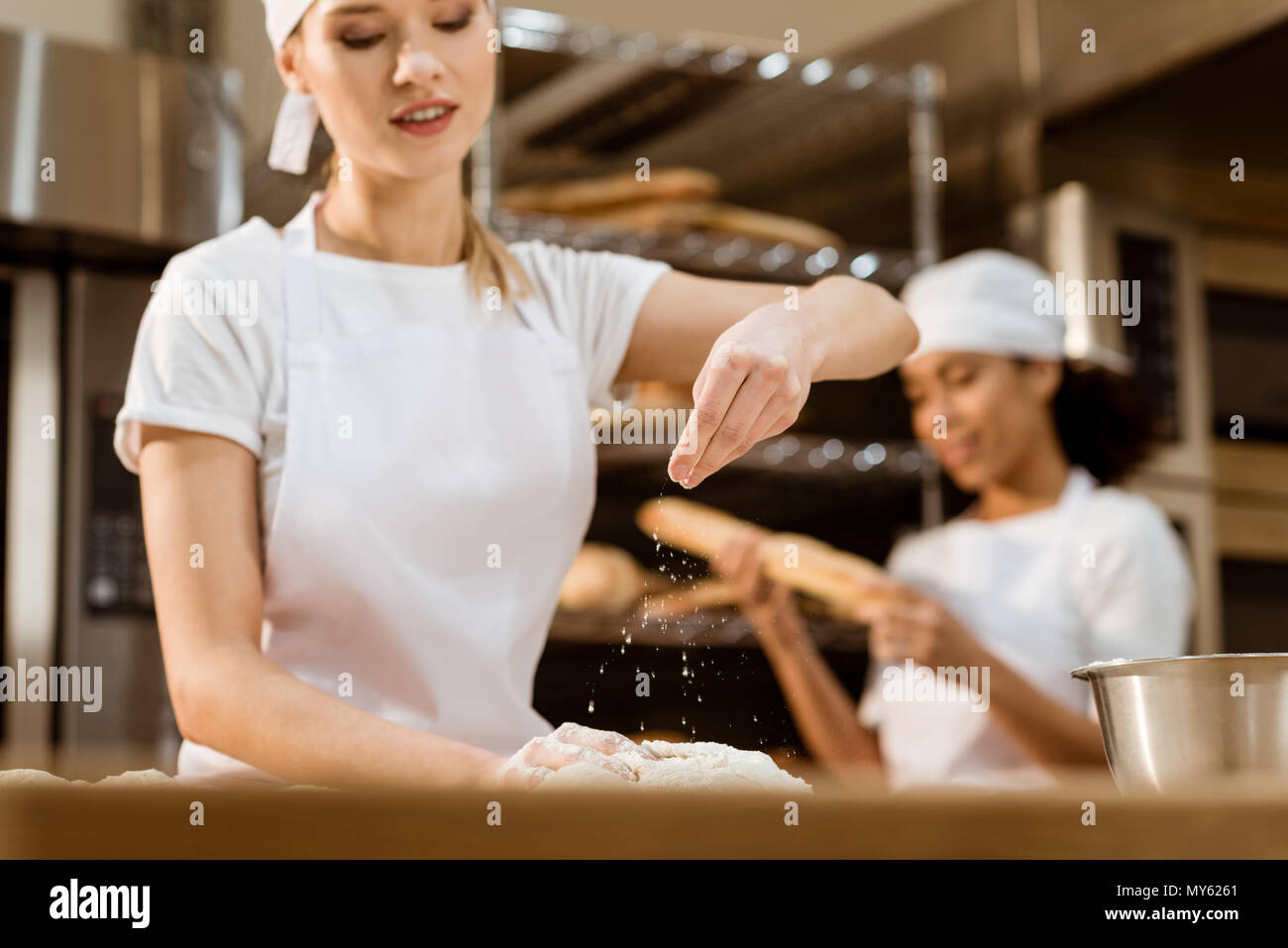 happy female baker kneading dough and pouring flour on it at baking ...
