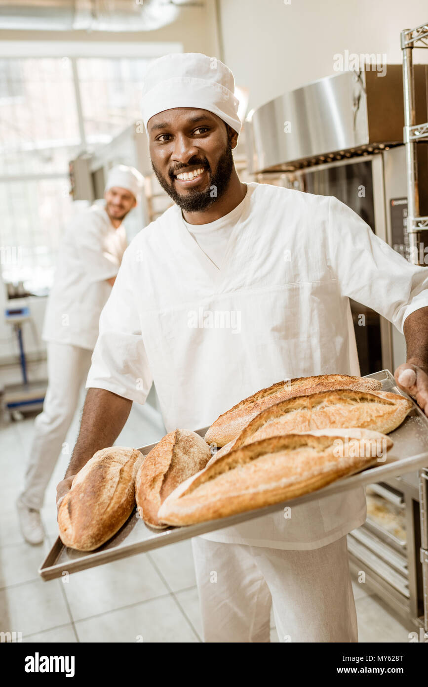 african american baker taking bread loaves from oven at baking ...