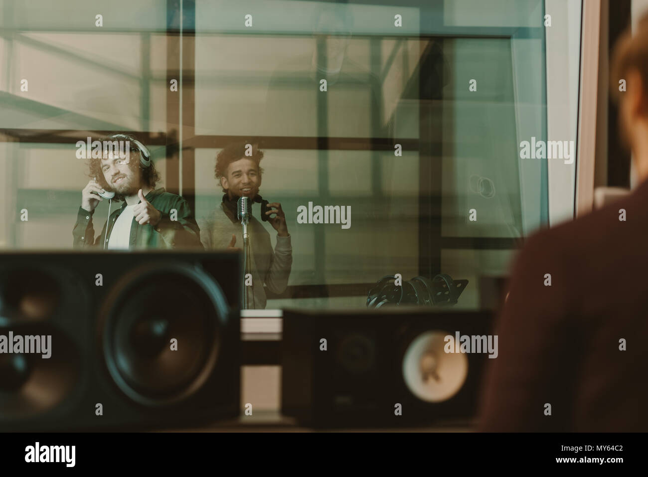 young happy musicians recording song behind glass at studio and showing ...