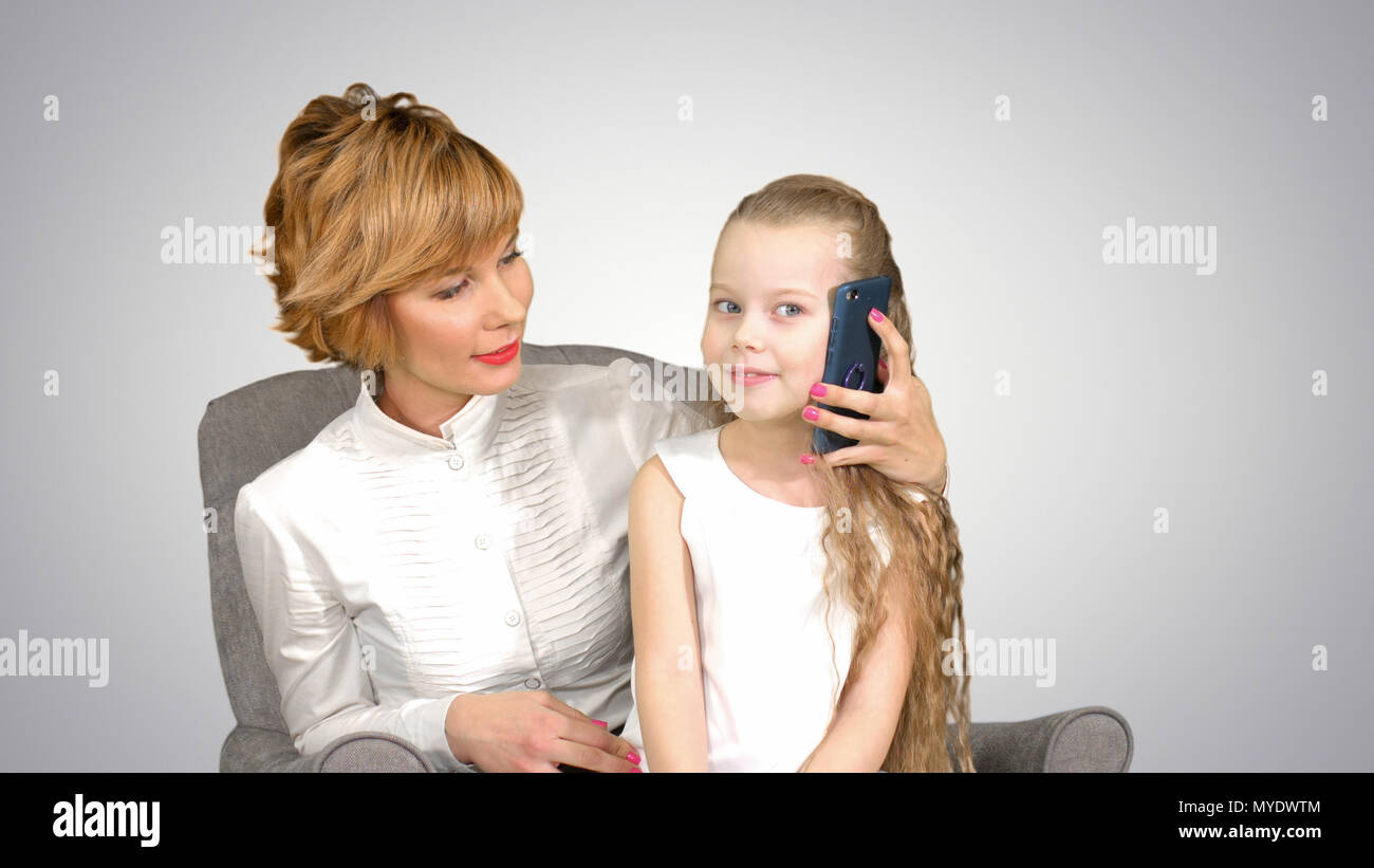 Little daughter having phone call, sitting near her mother on white ...