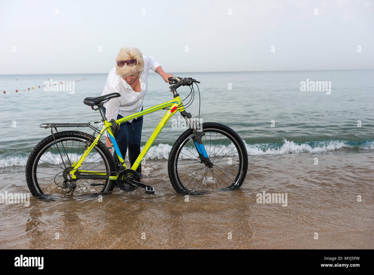 Woman in jeans standing in the water on a beach leaning over a bicycle ...
