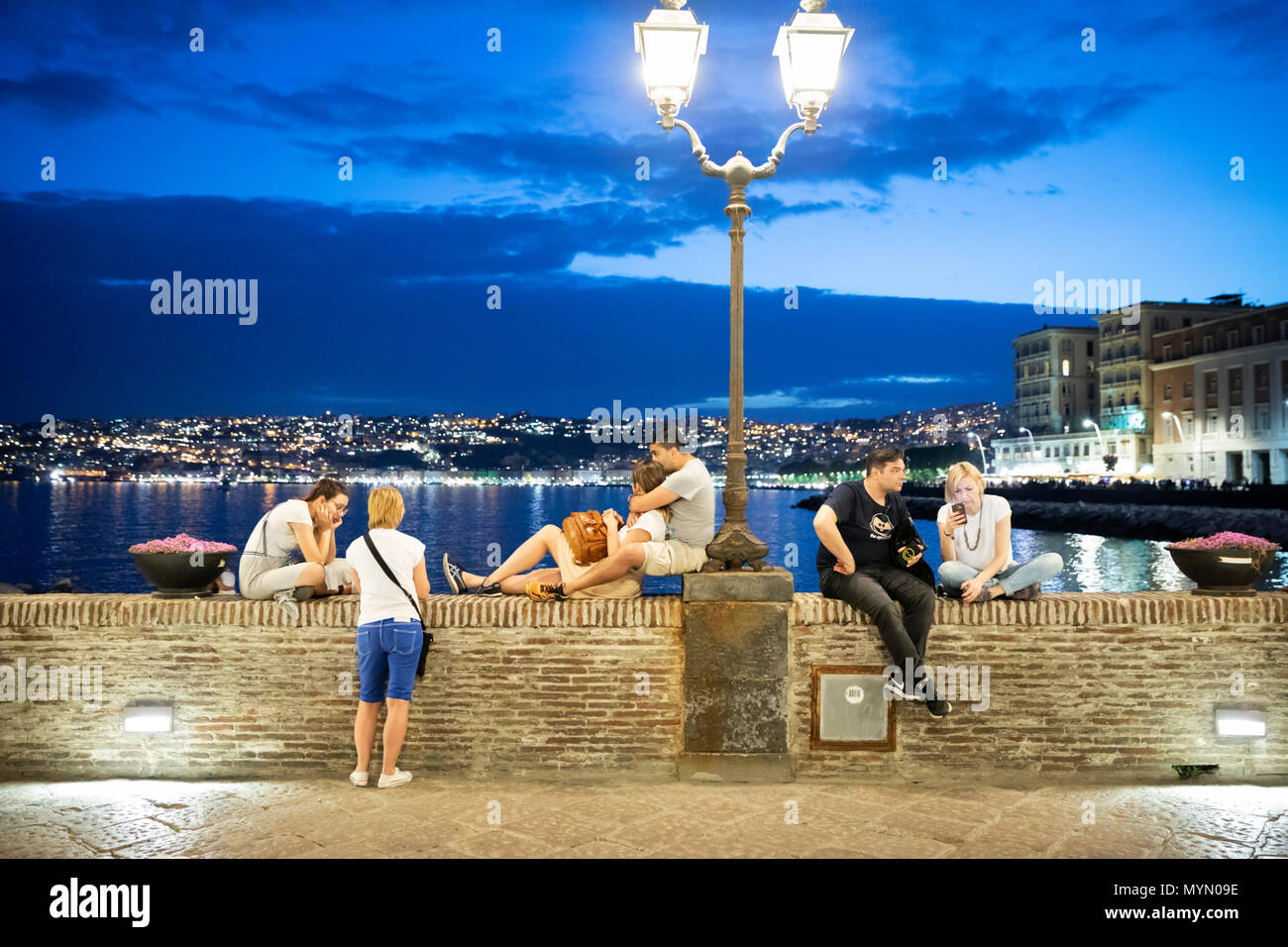 People sitting on wall under street lamp by the Castel dell Ovo at dusk ...