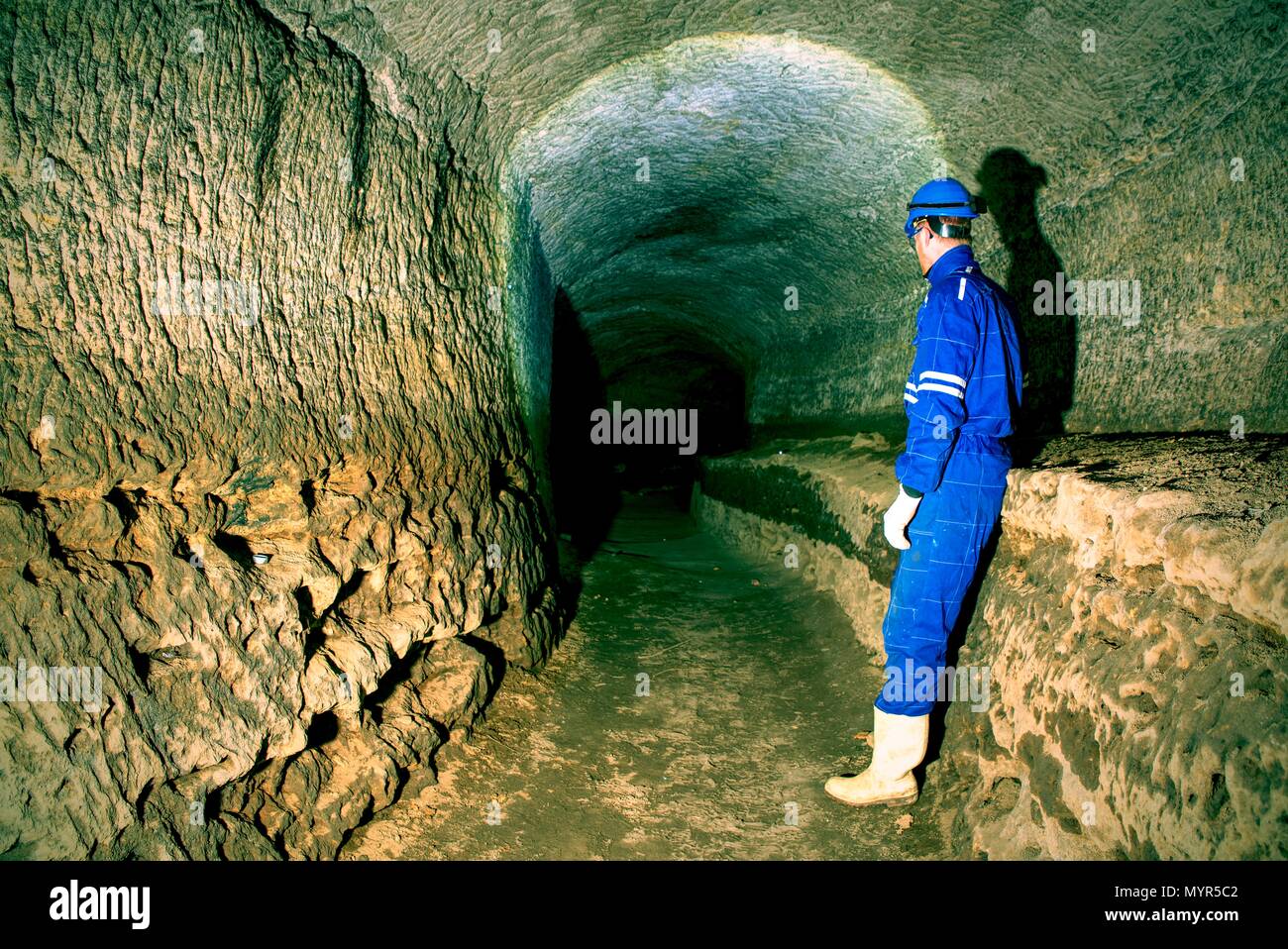 Underground work. Hunched worker in a blue overall and a safety helmet ...
