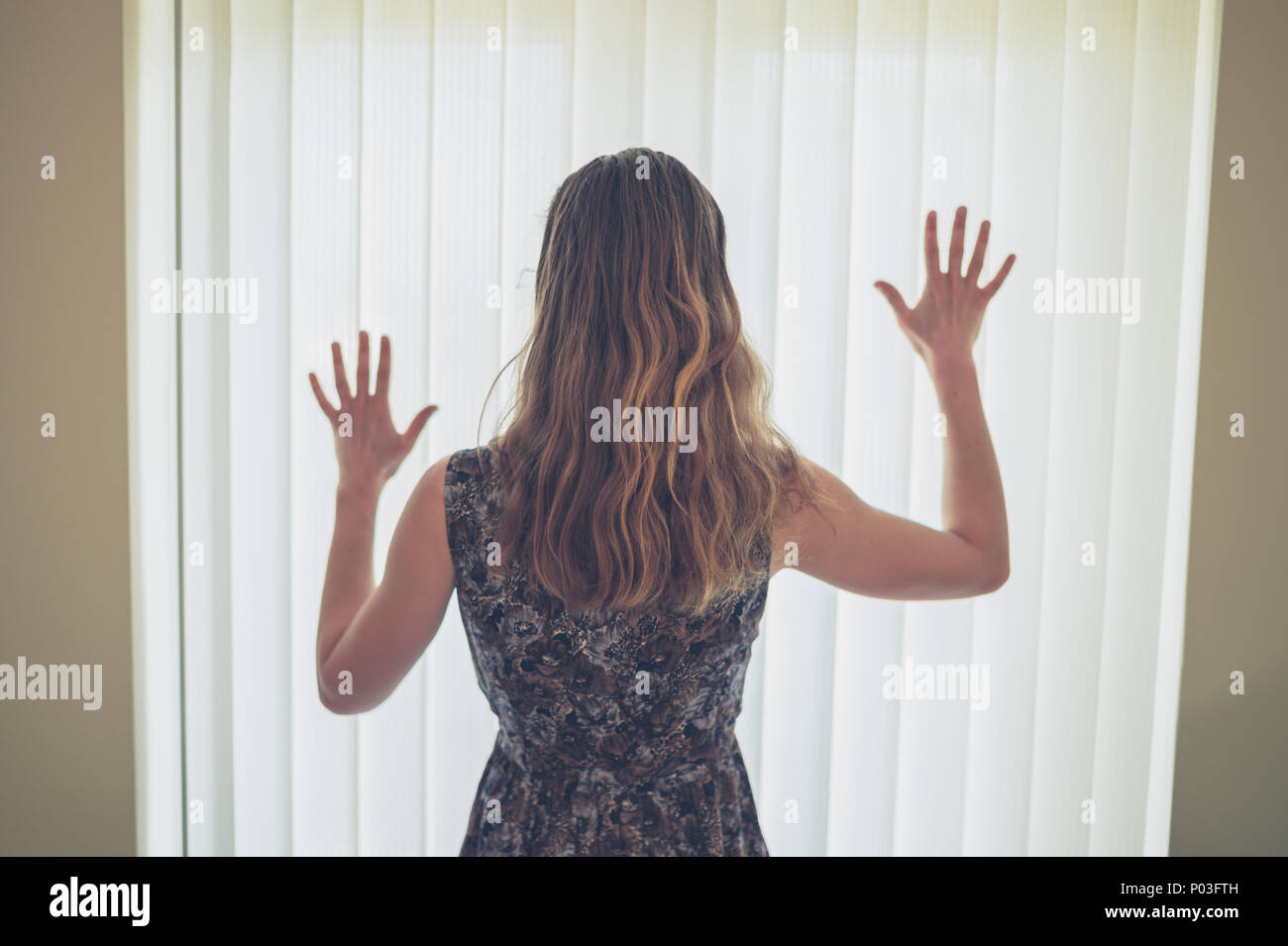 A young woman is standing by the window with her hands up against the ...