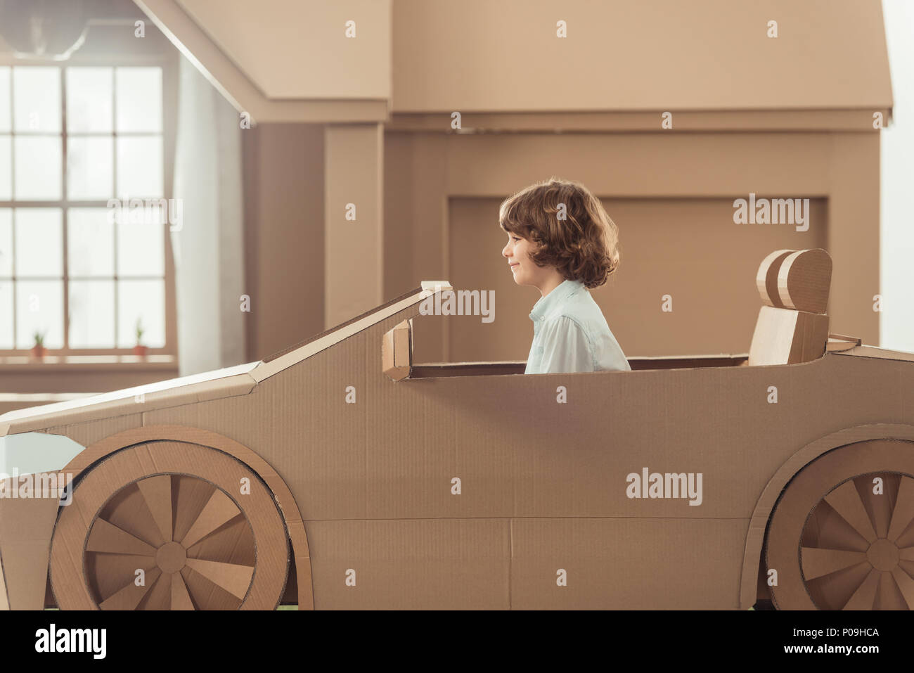 side view of little kid sitting in cardboard car in front of cardboard ...