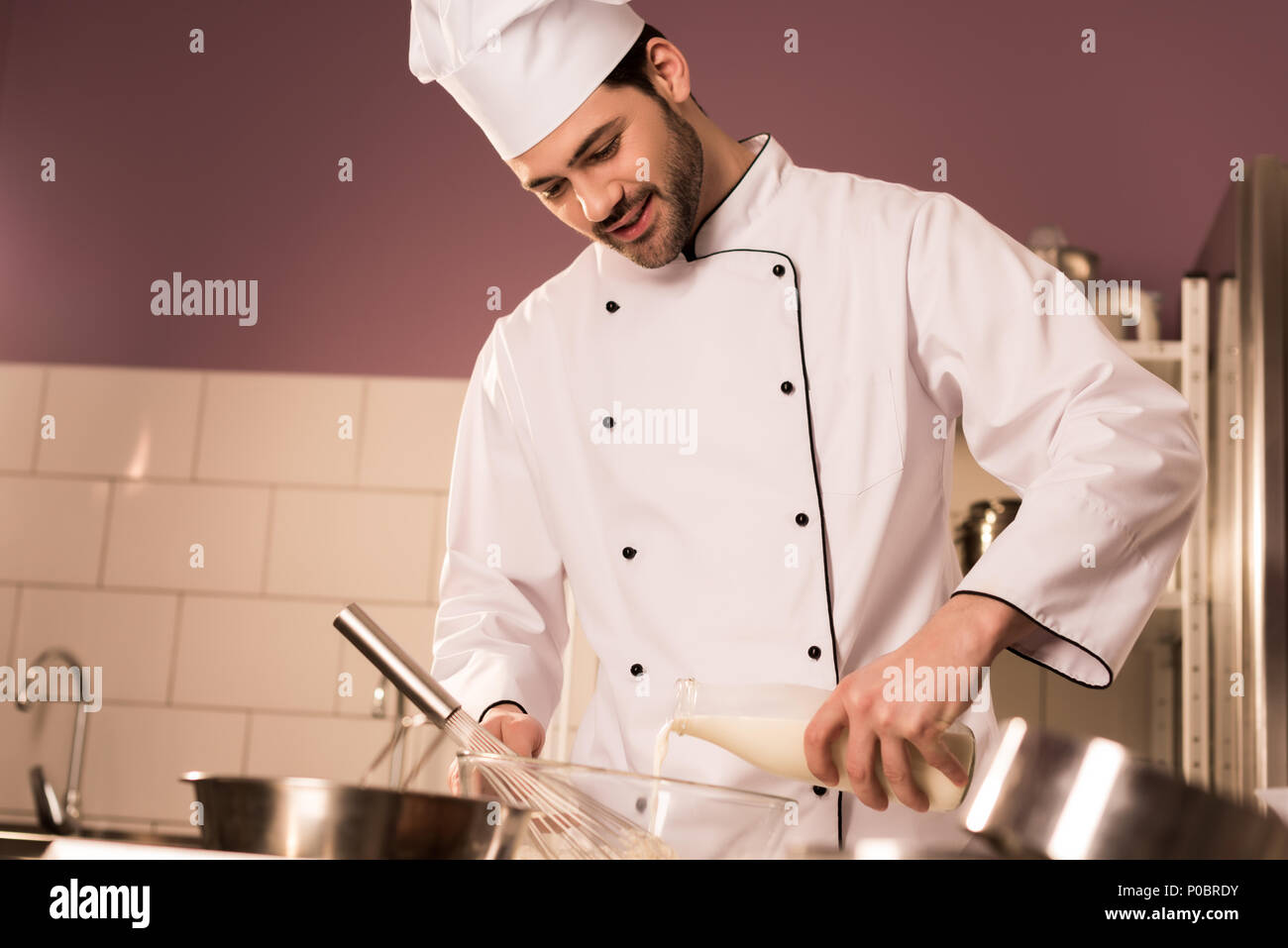 portrait of confectioner in chef hat pouring milk into bowl while ...