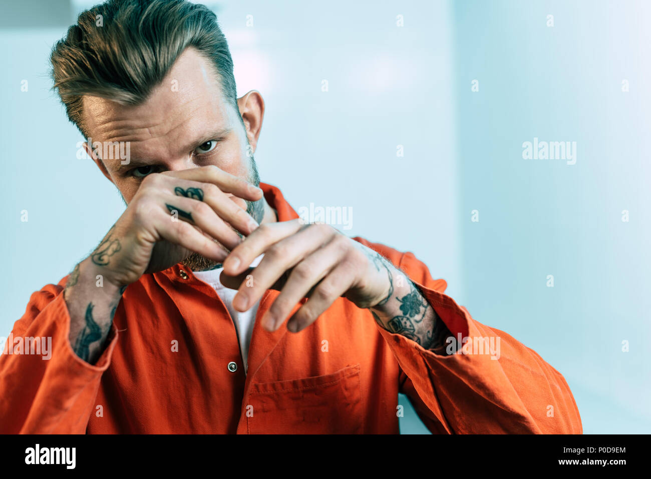 prisoner with tattoos covering face with hands and looking at camera ...
