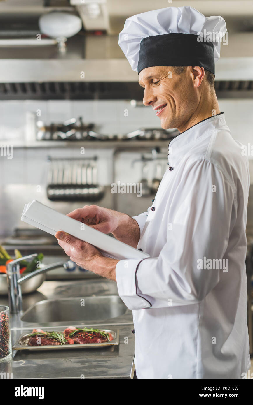 side view of handsome chef reading recipe book at restaurant kitchen ...