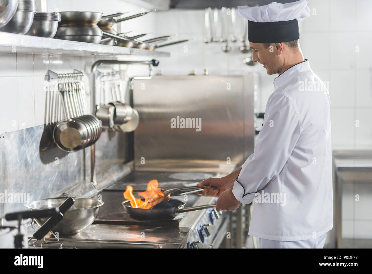 side view of handsome chef frying steak with fire at restaurant kitchen ...