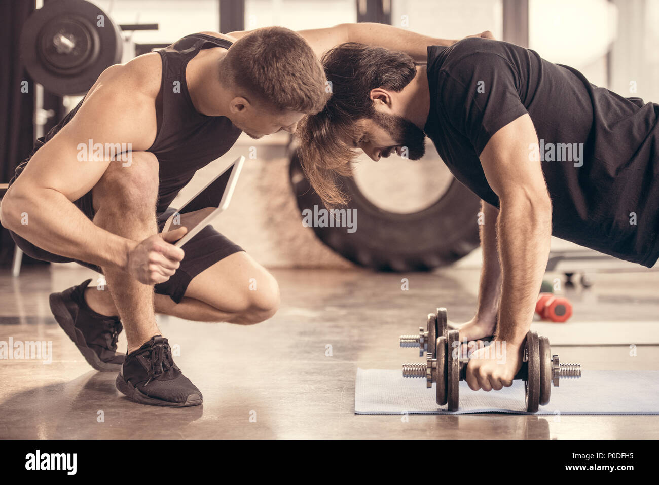 side view of muscular trainer pushing man doing push ups on dumbbells ...