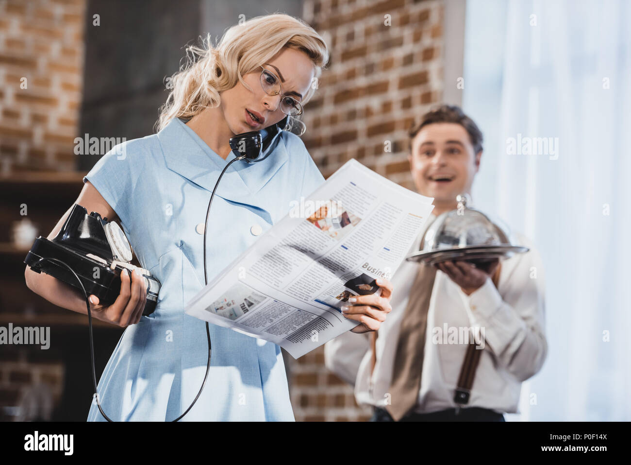 woman reading newspaper and talking by vintage telephone while husband ...
