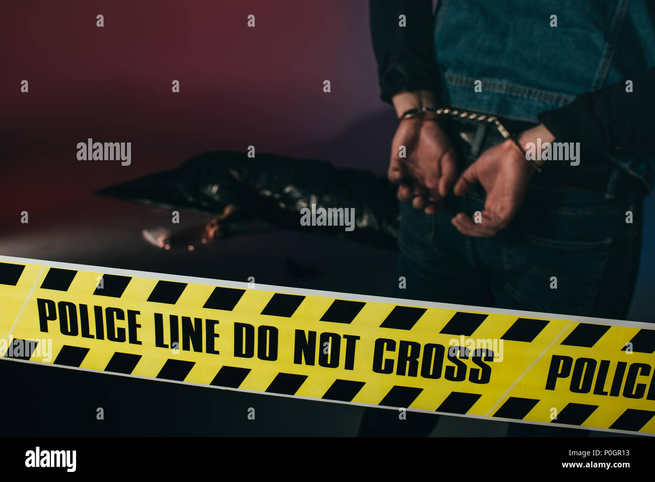 Rear view of man in cuffs by dead body behind yellow tape on dark ...