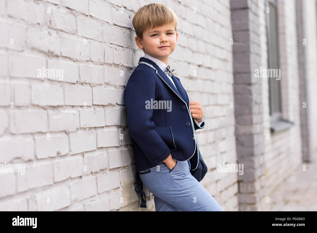 stylish boy with hand in pocket leaning at brick wall and looking at ...
