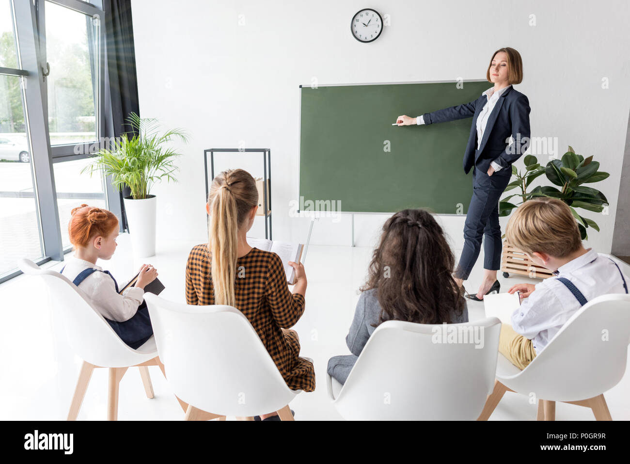 young female teacher holding chalk and pointing at blackboard while ...