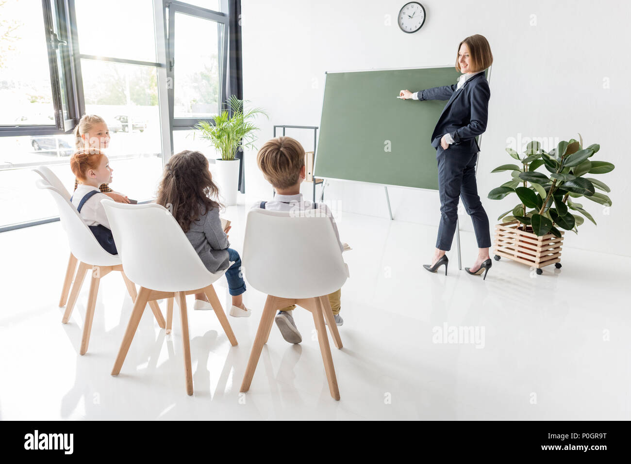 young female teacher holding chalk and pointing at blackboard while ...