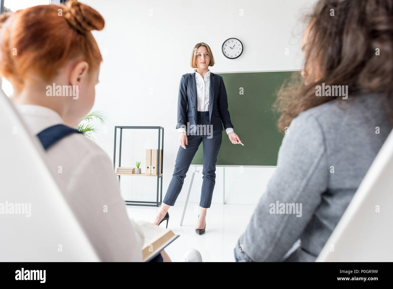 smiling young teacher holding chalk and looking at schoolkids during ...