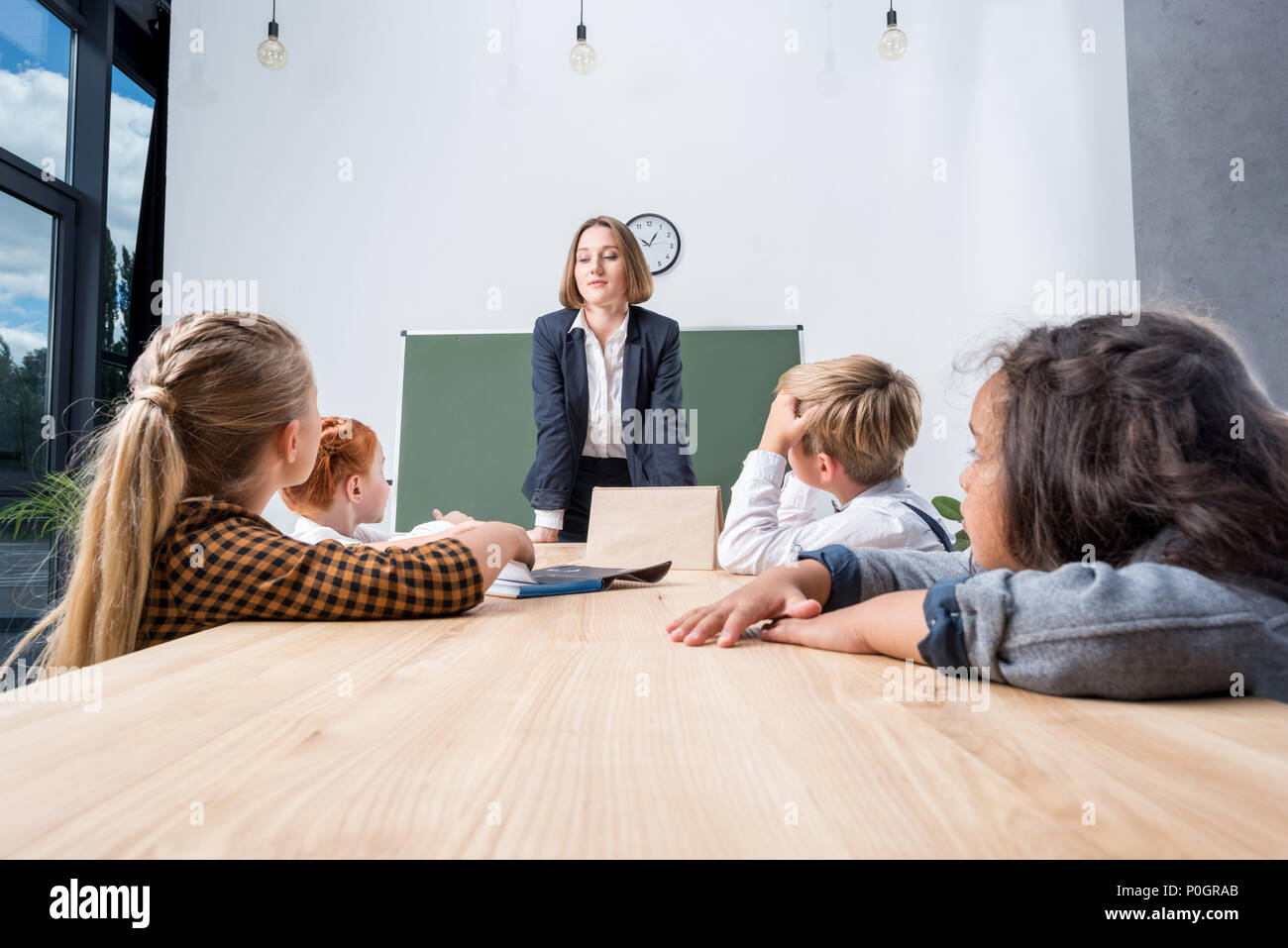 smiling young teacher leaning at desk and looking at cute multi ethnic ...