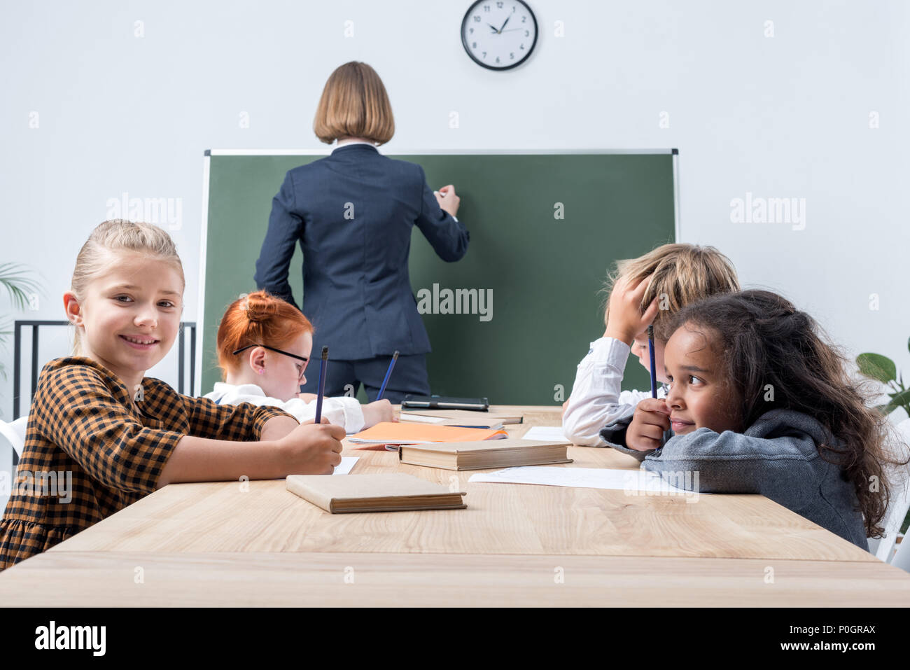 back view of teacher writing on chalkboard and smiling children ...