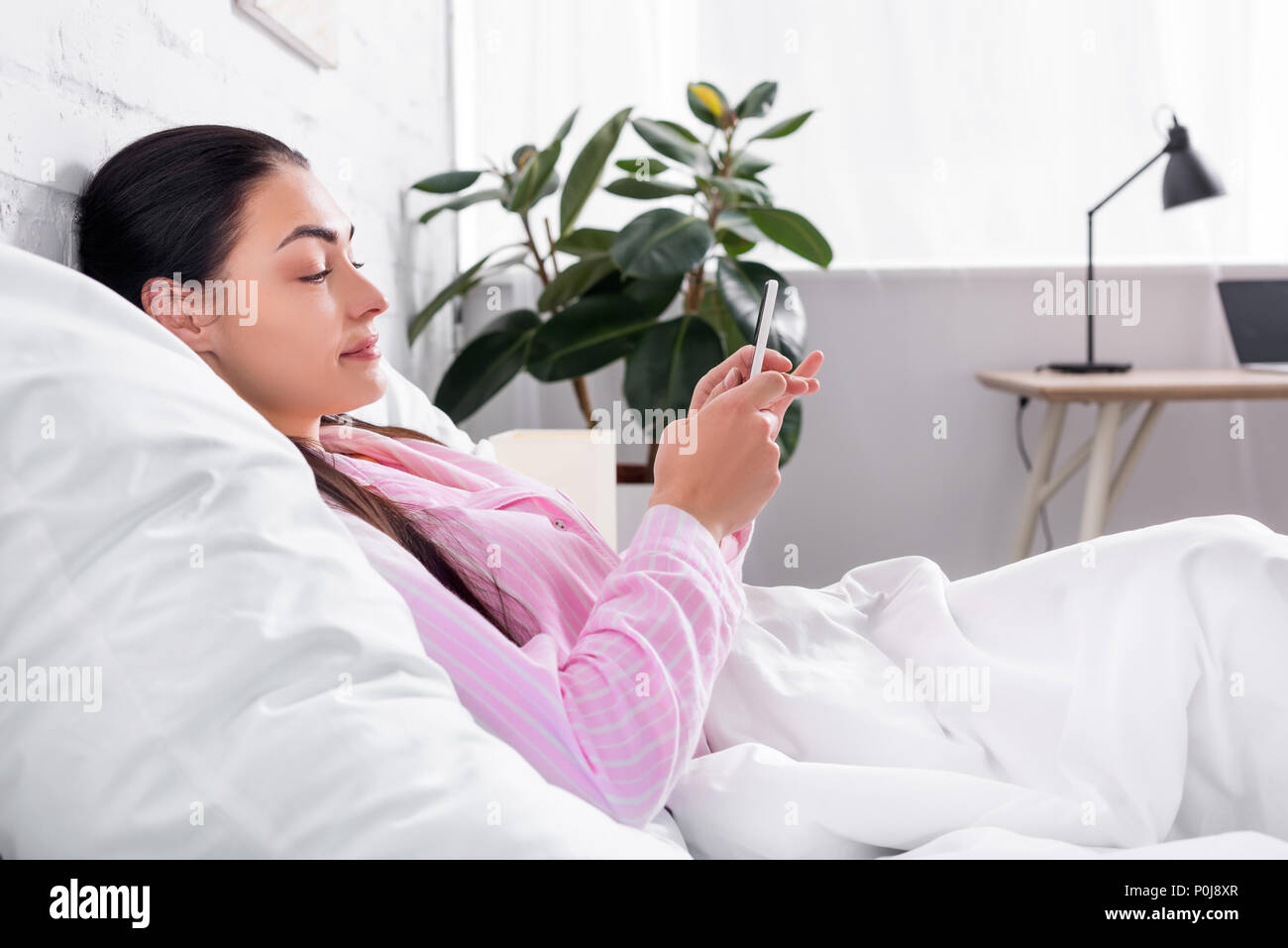 side view of attractive woman in pajamas using smartphone while resting ...