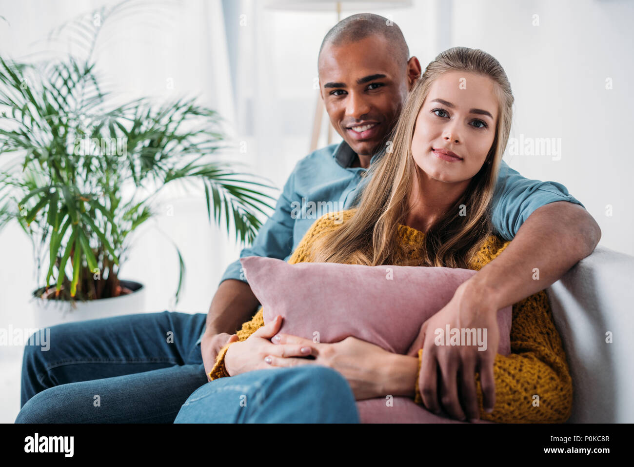 multicultural couple hugging on sofa and looking at camera Stock Photo ...