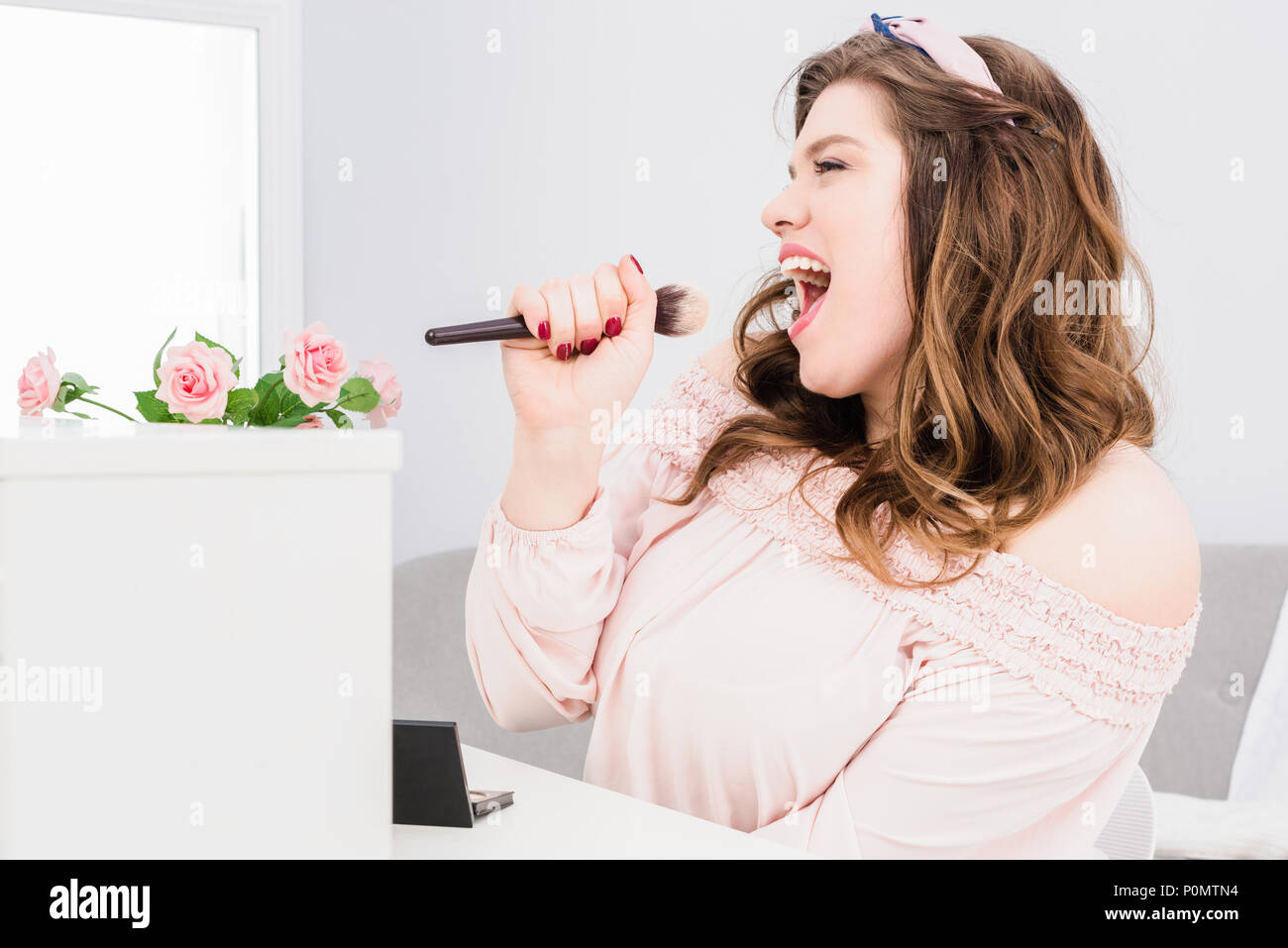 emotional young woman singing with makeup brush in hand as microphone ...
