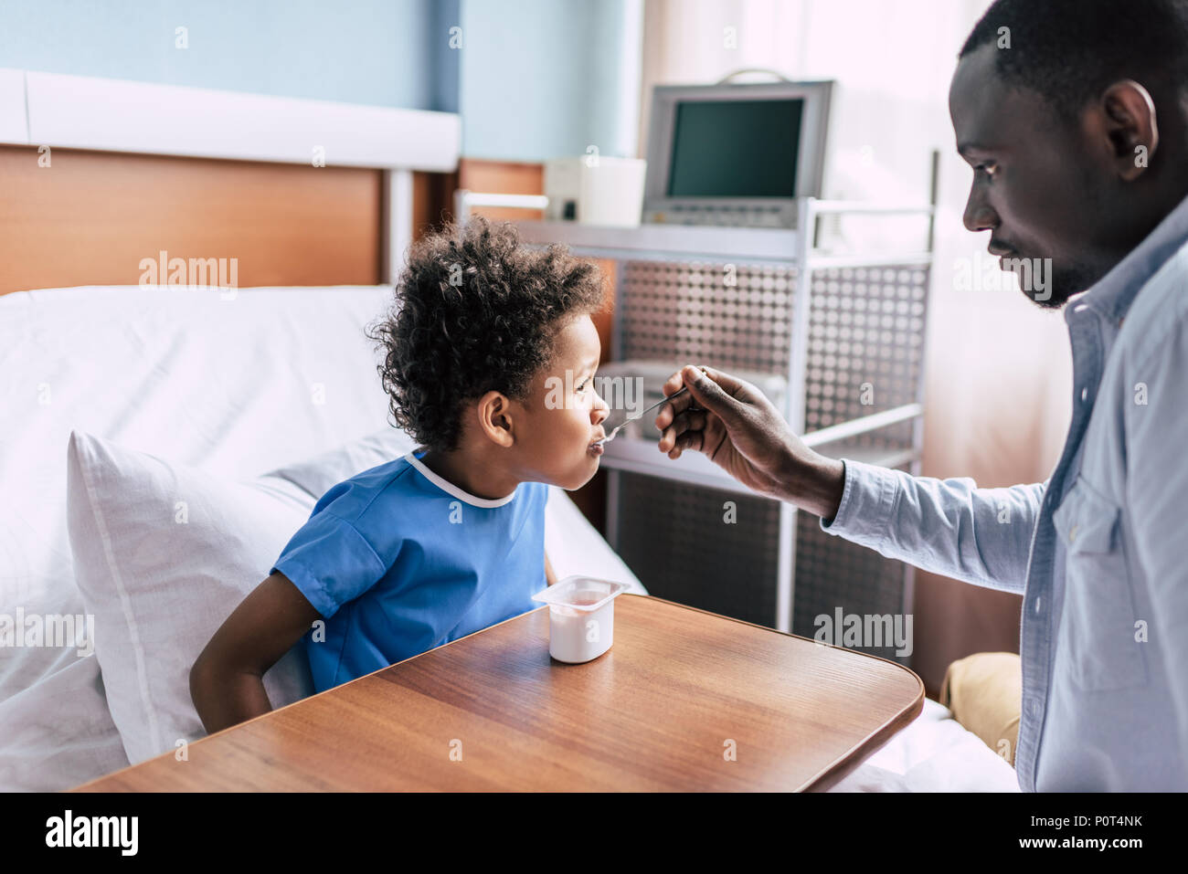 side view of african american father feeding little sick son in clinic ...