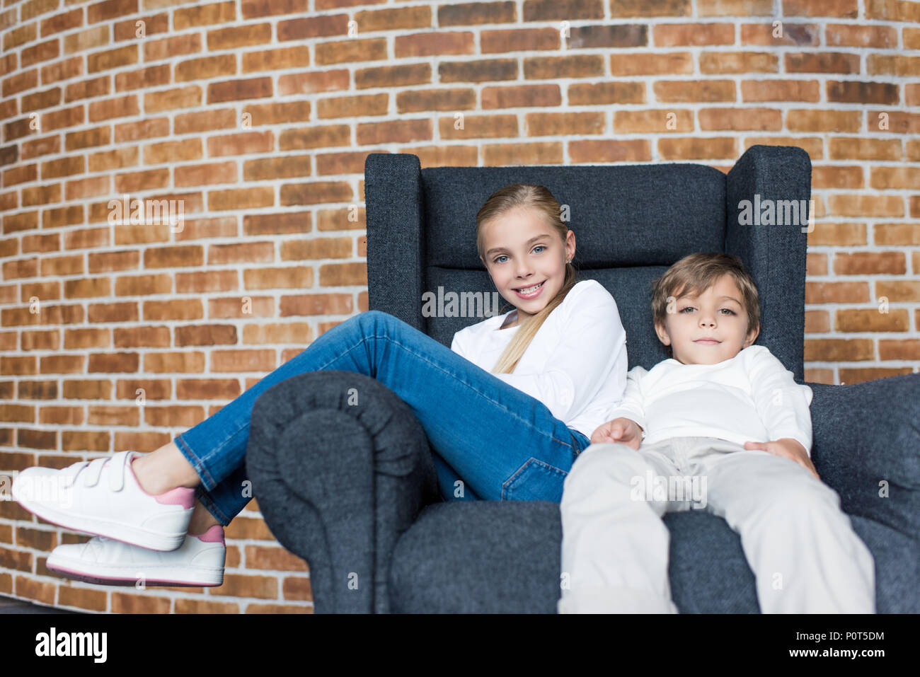 smiling siblings looking at camera while resting on armchair at home ...