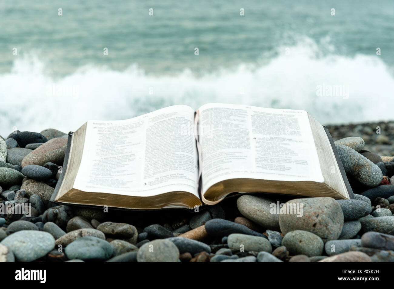 Bible open on top of small stones in front of the green sea with waves ...