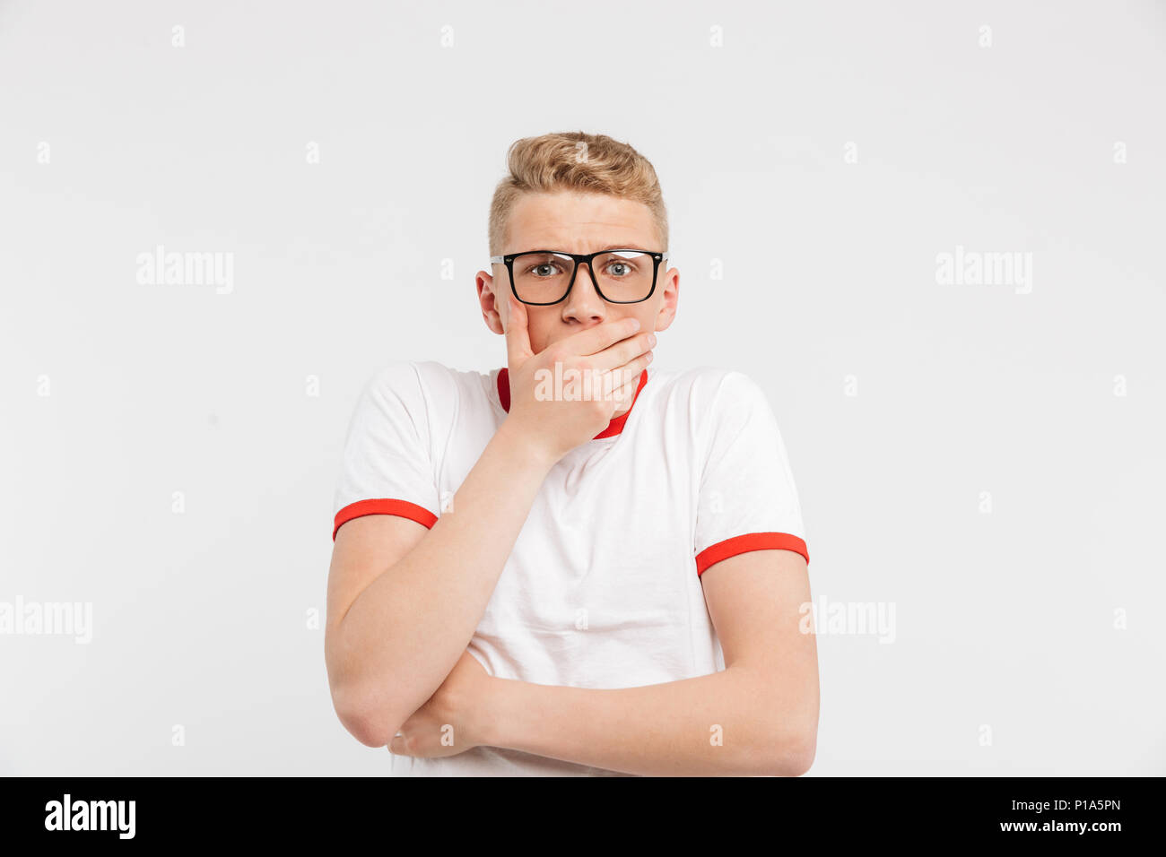 Portrait of a shocked teenage boy in eyeglasses covering face isolated ...