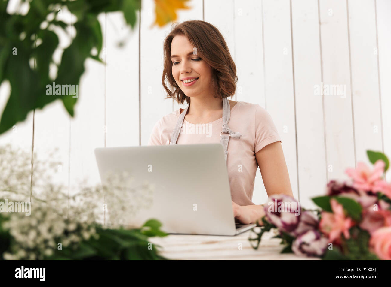 Young cute cheerful florist lady standing with flowers in workshop ...