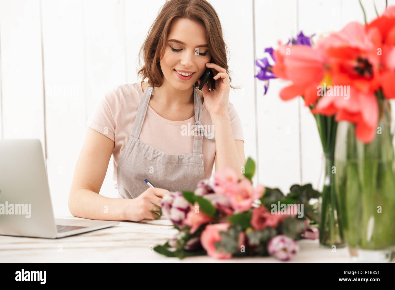 Young cute cheerful florist lady over flowers in workshop talking by ...
