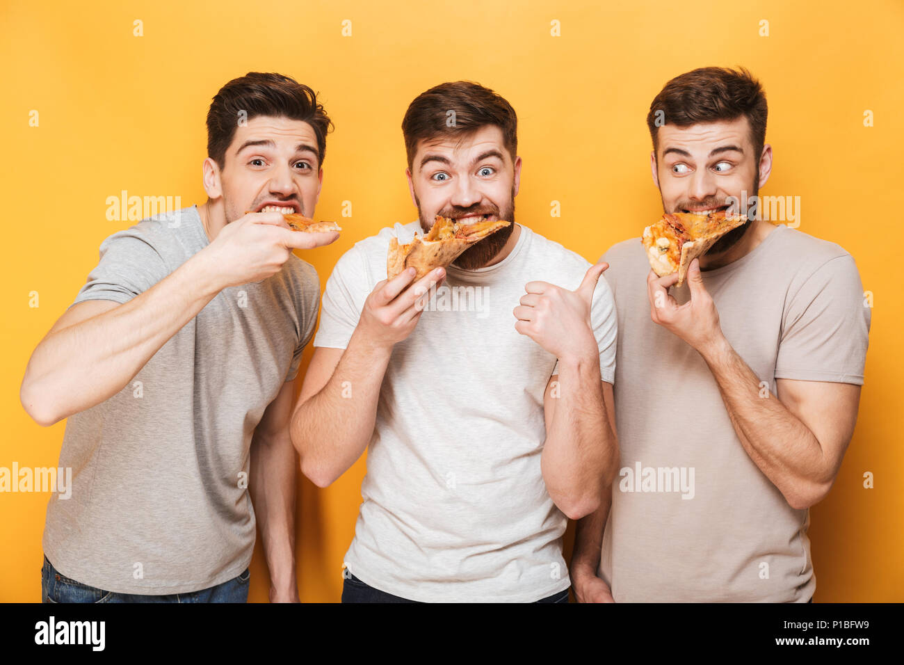 Three young happy men eating pizza isolated over yellow background ...